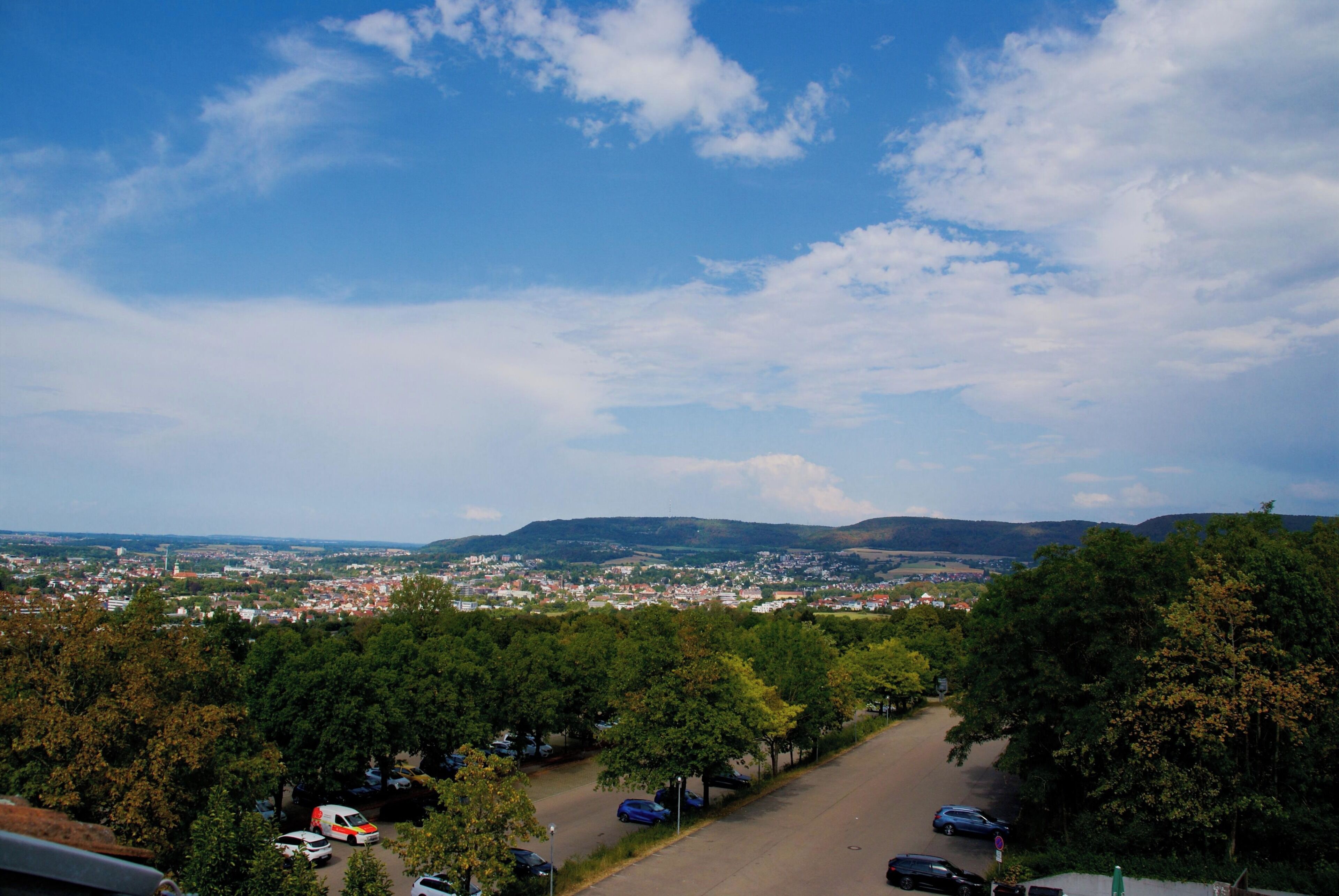 Paisagem urbana com vista para uma estrada arborizada e carros estacionados, que leva a uma área urbana densamente construída e uma cadeia montanhosa ao longe.