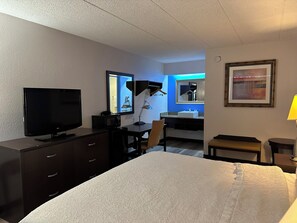 Quarto de hotel com cama, escrivaninha com computador, televisão, espelho, pia e quadro na parede.