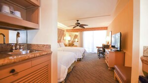 Quarto de hotel com cozinha compacta, duas camas, escrivaninha, televisão e ventoinha no teto.