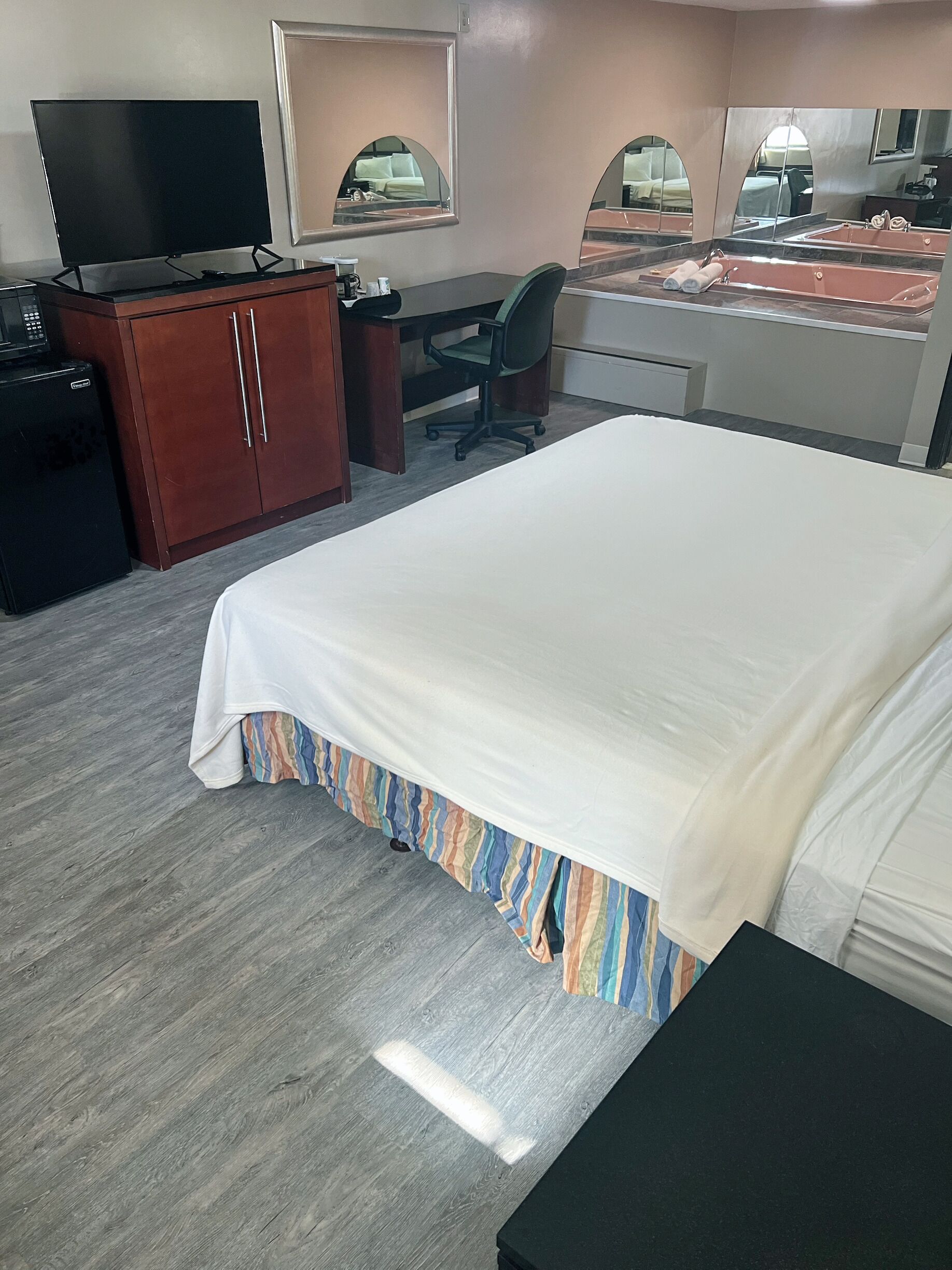 Quarto de hotel com cama, escrivaninha, cadeira, televisão, guarda-roupa e banheiro com banheira e espelho.