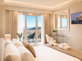 Quarto de hotel com cama, escrivaninha, cadeira, janela grande com vista para a cidade e varanda.
