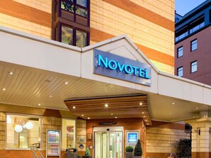 Exterior do hotel Novotel, com uma placa de destaque, entrada coberta e fachada de tijolos.