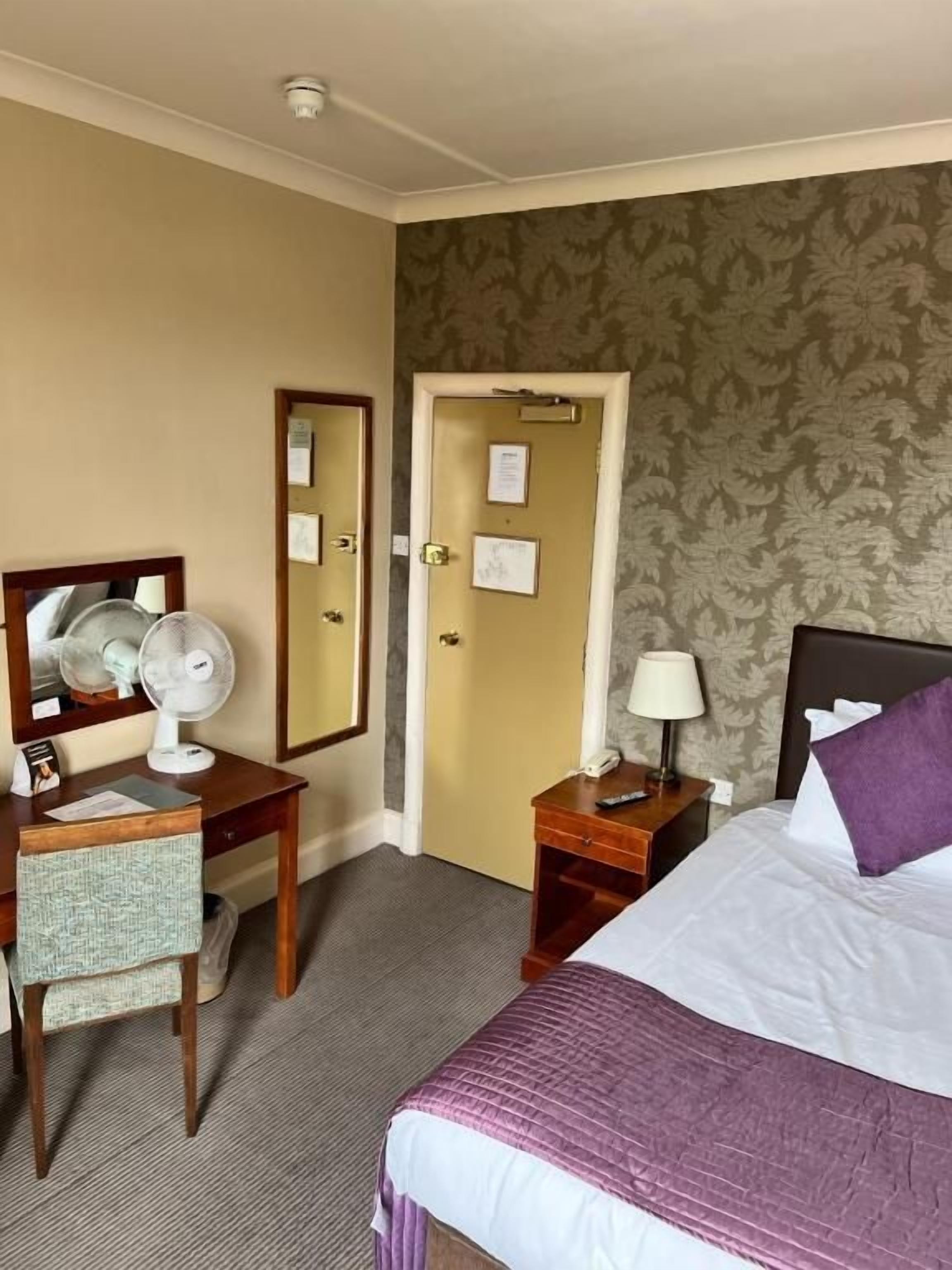 Quarto de hotel com cama, escrivaninha e cadeira, mesa de cabeceira com abajur, e porta com espelho.