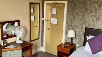 Quarto de hotel com cama, escrivaninha e cadeira, mesa de cabeceira com abajur, e porta com espelho.