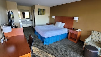 Quarto de hotel com cama, escrivaninha, cadeira, espelho e banheiro visível através de uma porta aberta.