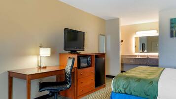 Quarto de hotel com cama, escrivaninha com cadeira, televisão, frigorífico, micro-ondas e casa de banho com espelho e lavatório.