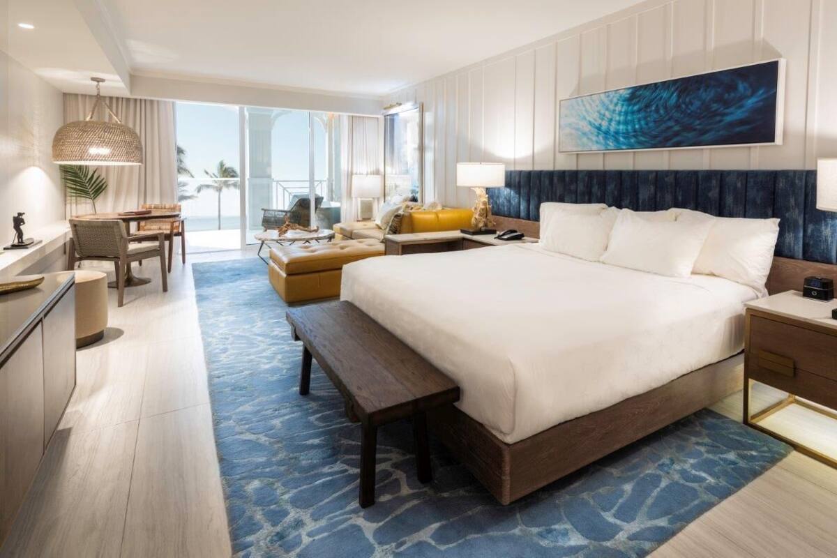 Quarto de hotel moderno com uma cama grande, uma escrivaninha, uma cadeira, um banco e vista para o oceano.