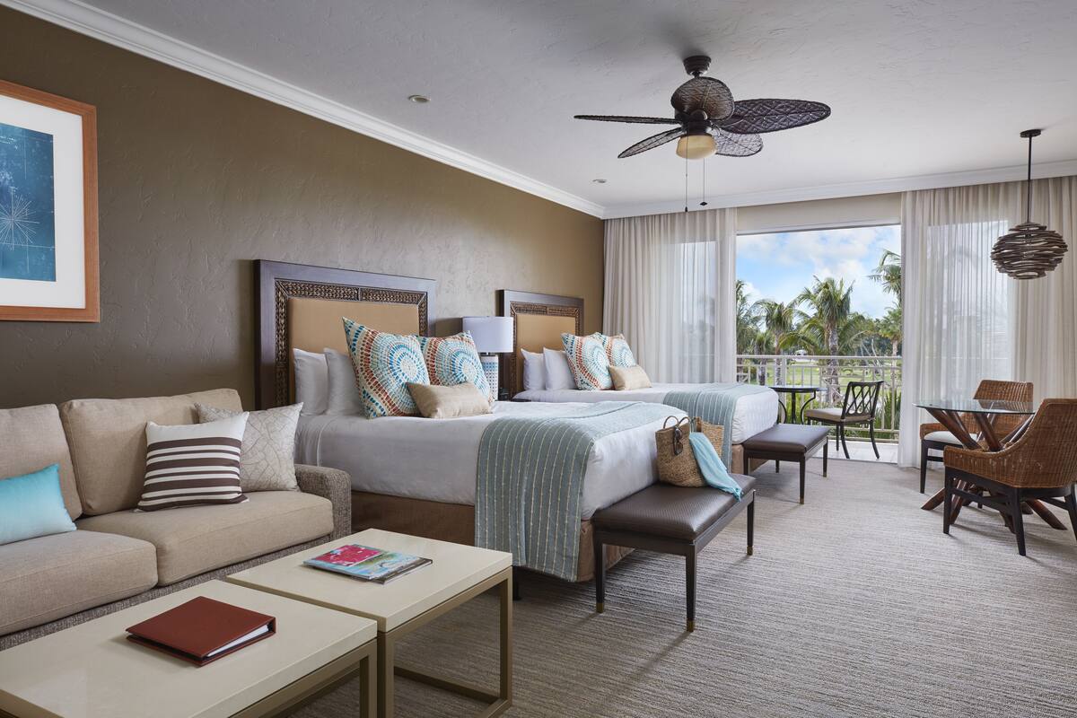 Quarto de hotel com duas camas, um sofá, uma mesa de jantar e varanda com cadeiras.