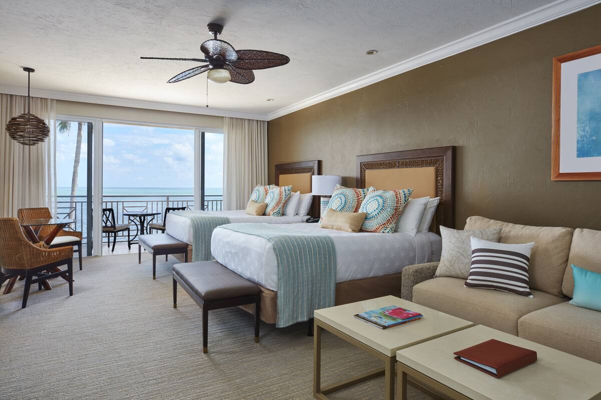 Quarto de hotel com duas camas, um sofá, uma mesa de jantar e uma varanda vista para o oceano.