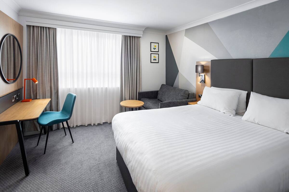 Quarto de hotel com uma cama grande, uma escrivaninha, uma cadeira, um sofá e uma mesa pequena. Há cortinas, um abajur e duas quadros na parede.