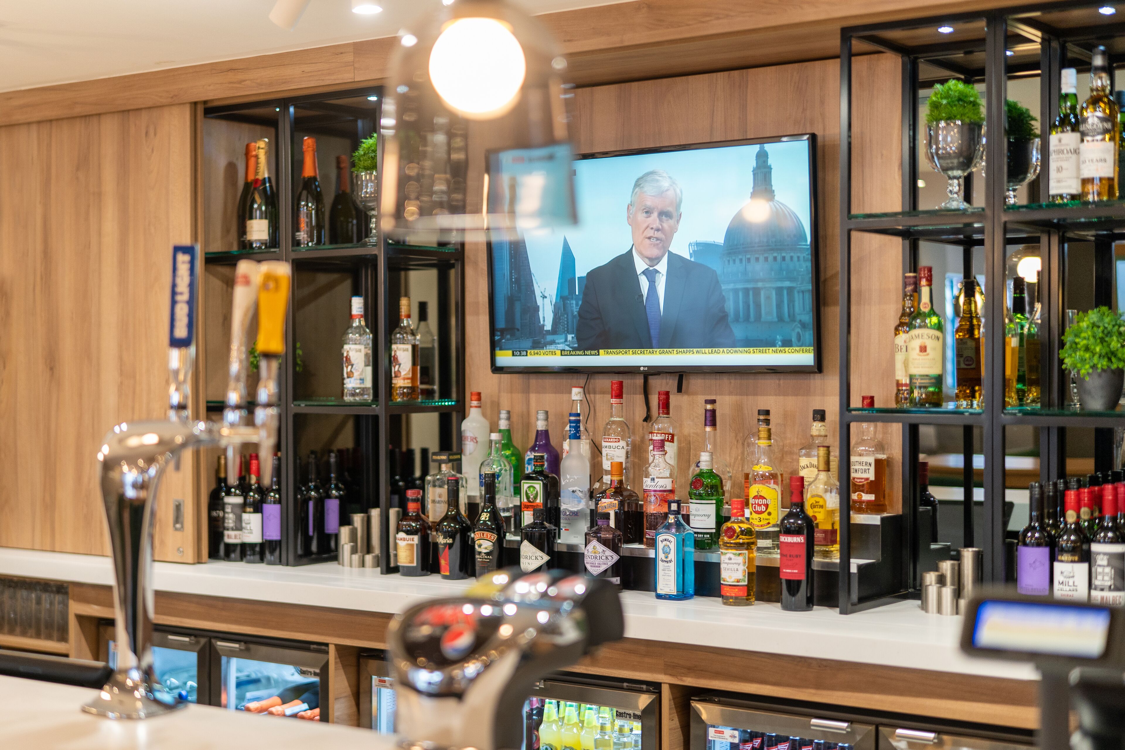 Uma área de bar com uma televisão exibindo uma notícia, prateleiras com diversas garrafas de bebidas e um balcão com torneira e copos.