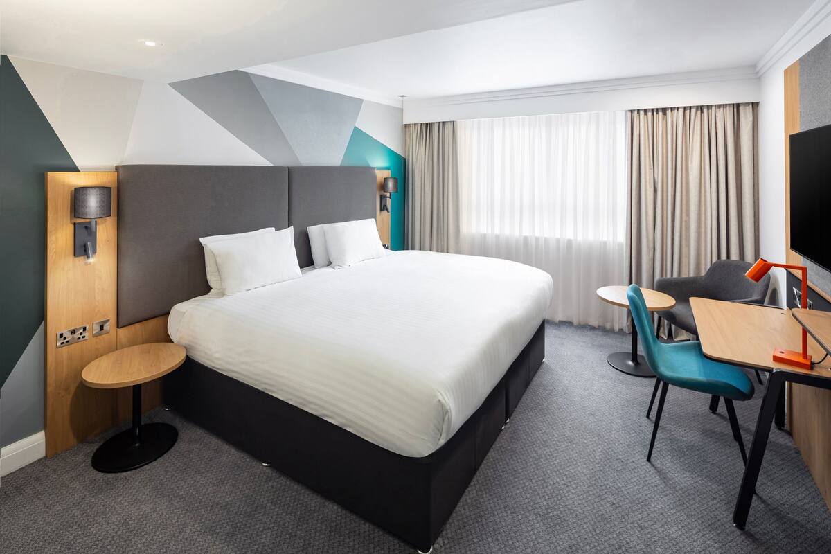 Quarto de hotel moderno com uma cama grande, uma escrivaninha com cadeira, uma televisão e uma mesa redonda pequena.