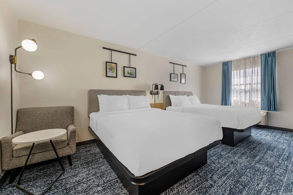 Quarto de hotel com duas camas, uma cadeira, uma mesa e um abajur.