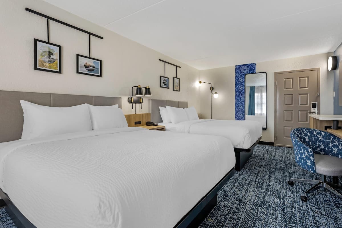 Quarto de hotel com duas camas, uma escrivaninha, uma cadeira e porta de banheiro.