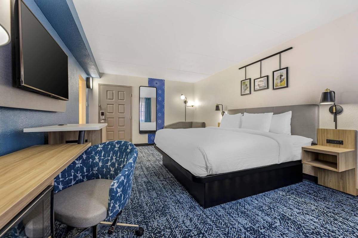 Quarto de hotel com cama, escrivaninha, cadeira, televisão, abajures e prateleira na parede.
