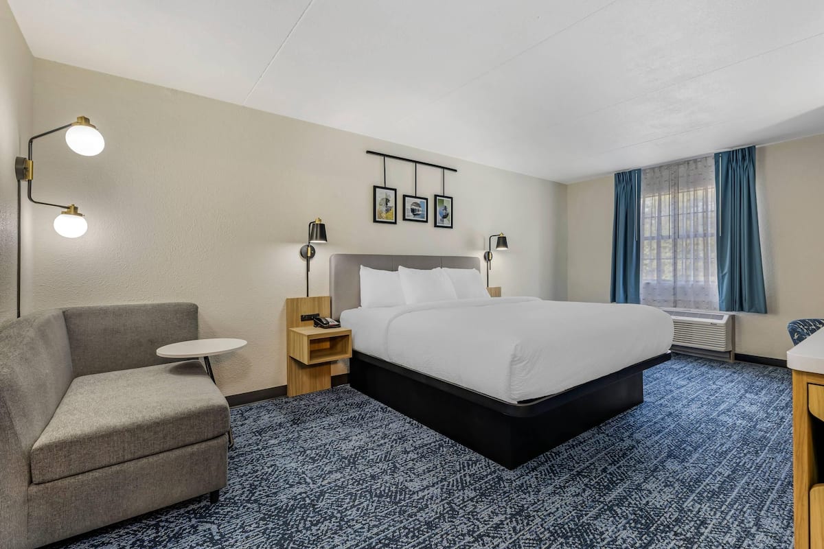 Quarto de hotel com cama, cadeira, mesas de cabeceira, luminárias e janela com cortinas.