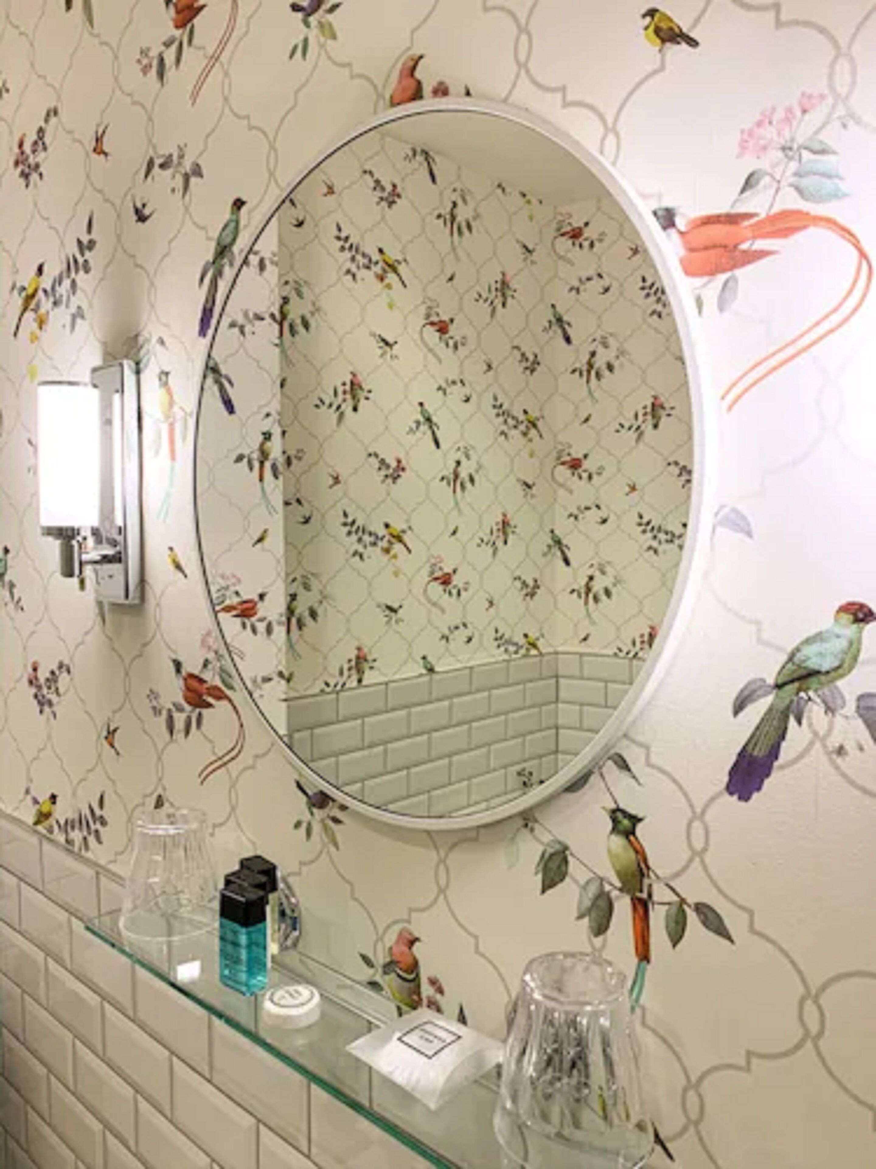 Casa de banho com espelho redondo, papel de parede floral e prateleira de vidro com artigos de higiene pessoal.