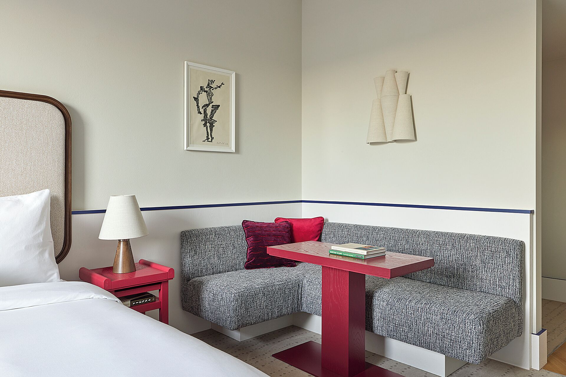 Quarto de hotel com uma cama, mesa de cabeceira vermelha, uma mesa com livros, um sofá cinza estampado e uma obra de arte emoldurada na parede.