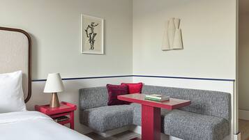 Quarto de hotel com uma cama, mesa de cabeceira vermelha, uma mesa com livros, um sofá cinza estampado e uma obra de arte emoldurada na parede.