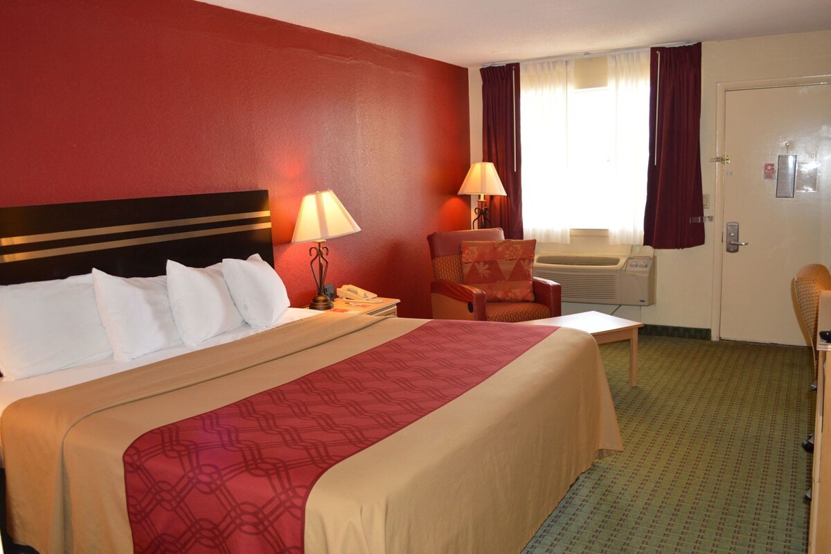 Quarto de hotel com cama, duas mesas de cabeceira, uma cadeira, escrivaninha, luminária, ar condicionado e janela com cortinas.