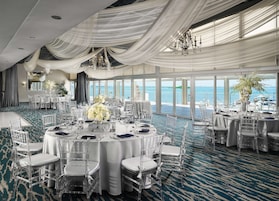 Uma sala de banquetes com mesas redondas, cadeiras transparentes e vista para o mar por grandes janelas.