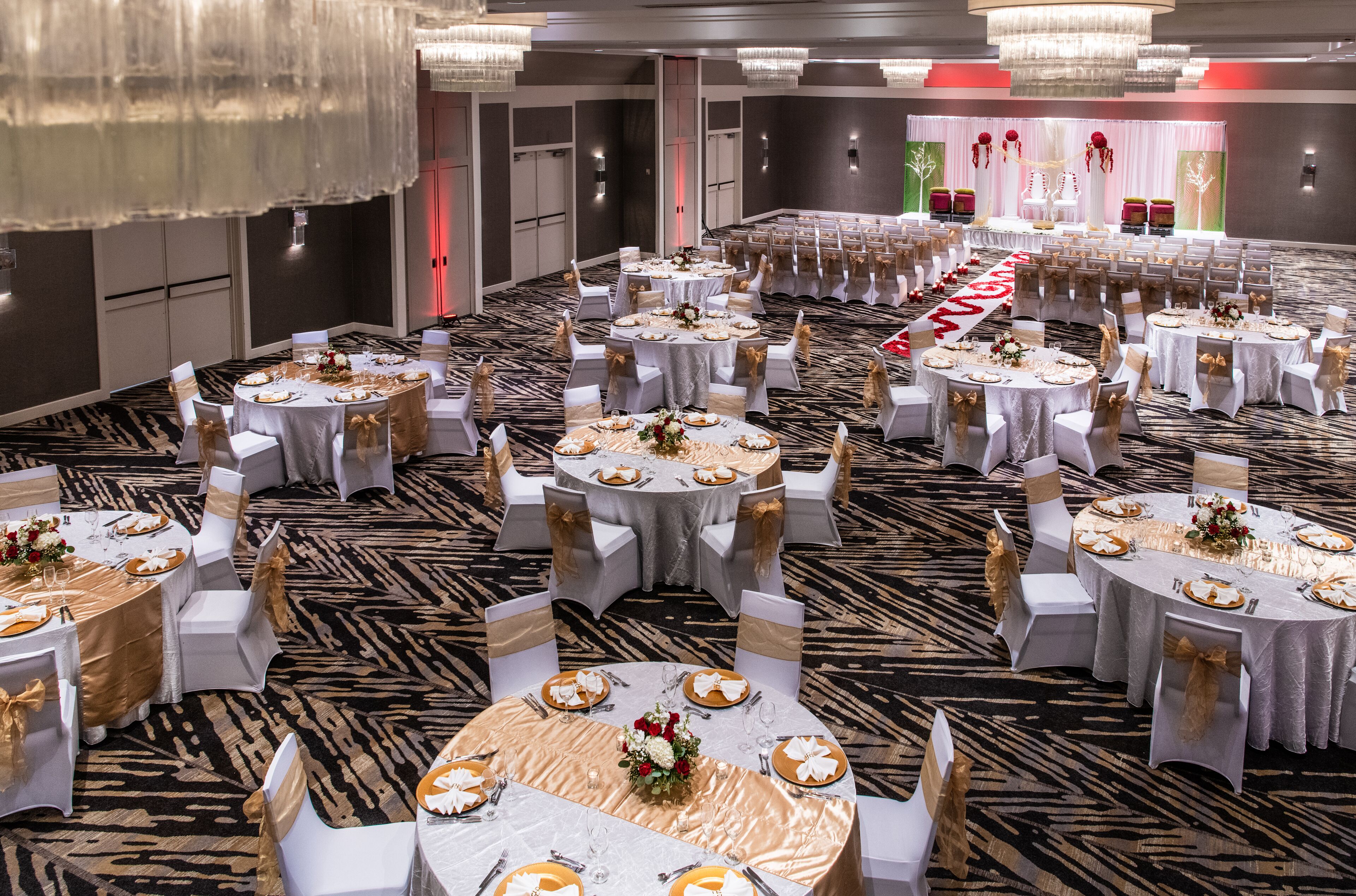 Uma sala de banquetes com mesas redondas dispostas para um evento formal, com toalhas brancas, fitas douradas e arranjos florais.