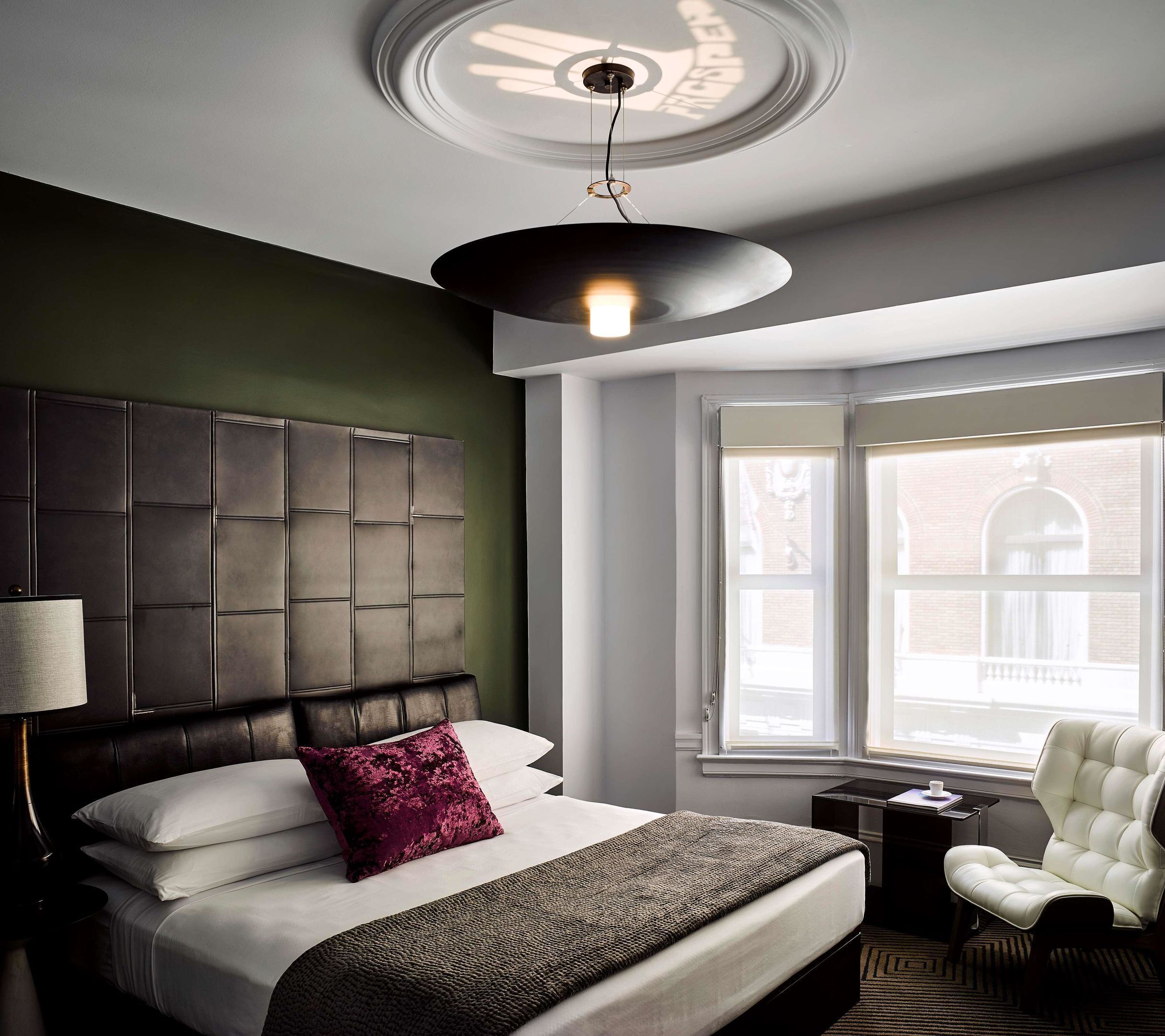 Quarto de hotel moderno com uma cama grande, cabeceira, janela com vista, uma cadeira e luminária no teto.