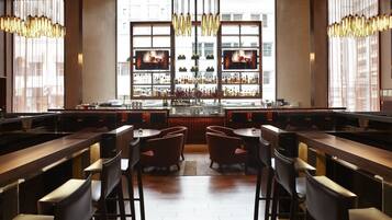Um bar com mesas e cadeiras de madeira, uma janela grande com cortinas e uma variedade de garrafas expostas.