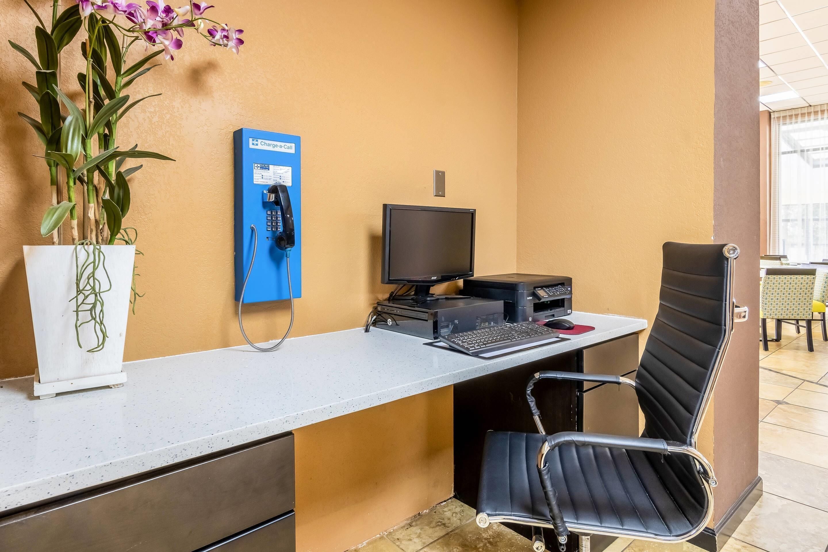 Uma mesa com computador, telefone e uma cadeira em um quarto com parede bege e planta em vaso.