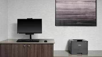 Um escritório moderno com um computador de mesa, teclado e rato sobre uma escrivaninha de madeira. Uma impressora Brother está no chão, ao lado da mesa. Um quadro abstrato emoldurado está na parede.