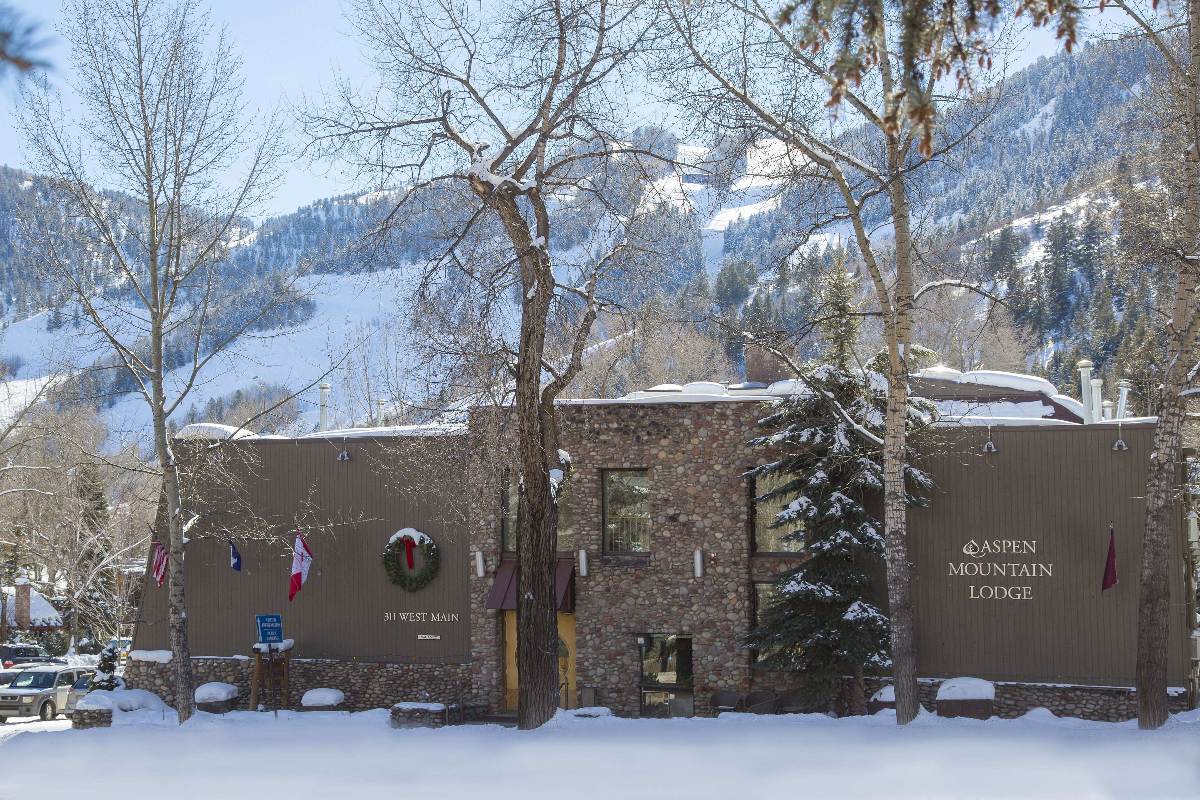 Uma pousada com fachada de pedra, uma placa com a inscrição "Aspen Mountain Lodge" e vista para montanhas cobertas de neve ao fundo.