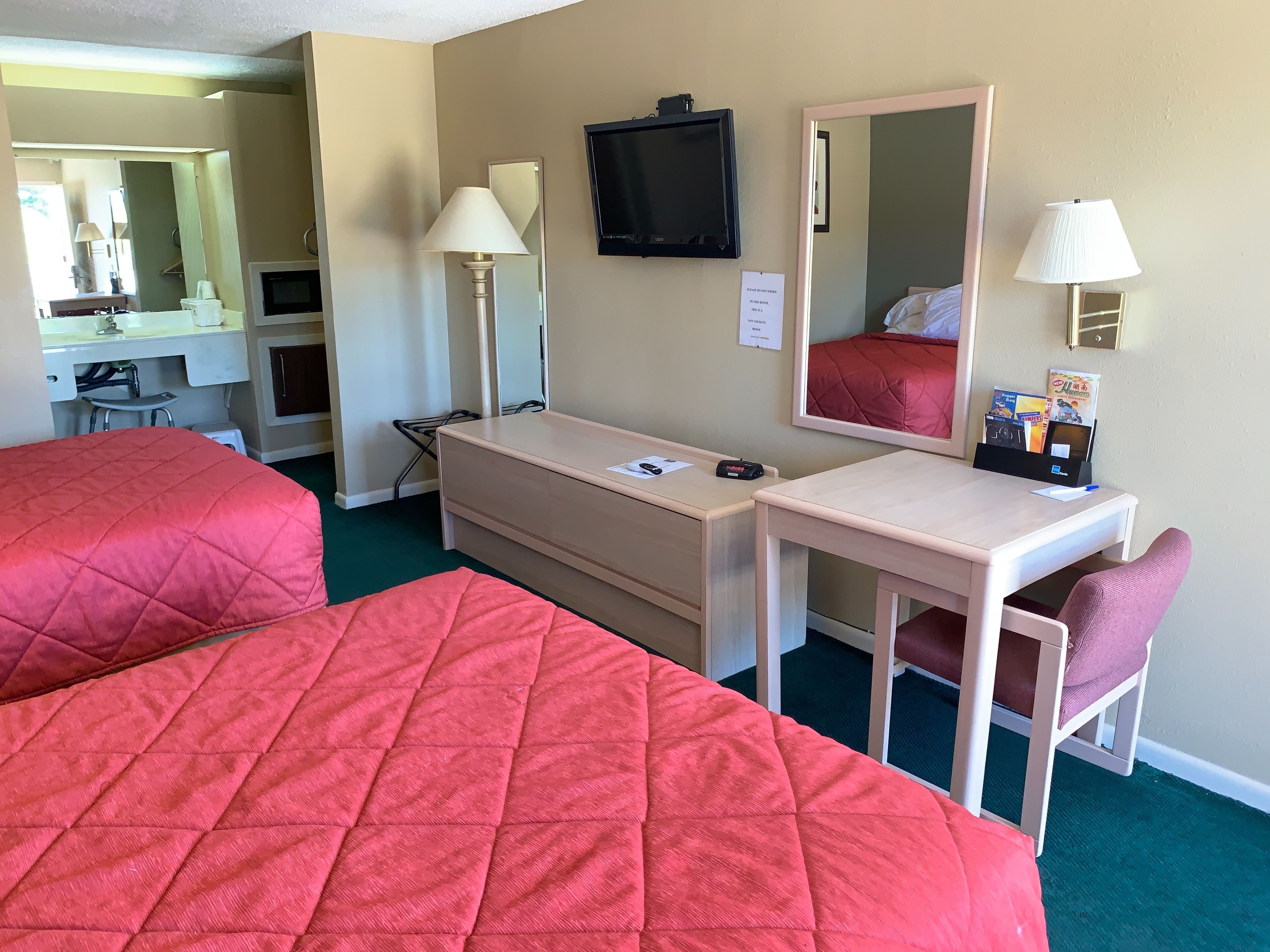 Quarto de hotel com duas camas, uma escrivaninha, uma cadeira, televisão, espelho e abajur.