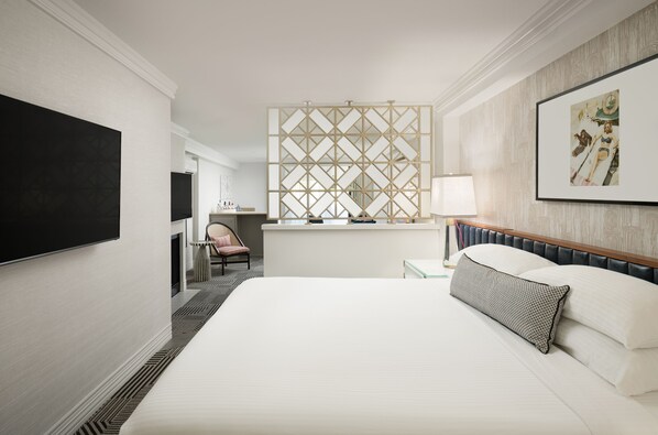 Quarto de hotel com uma cama grande, uma TV na parede, uma escrivaninha com abajur e um biombô decorativo.