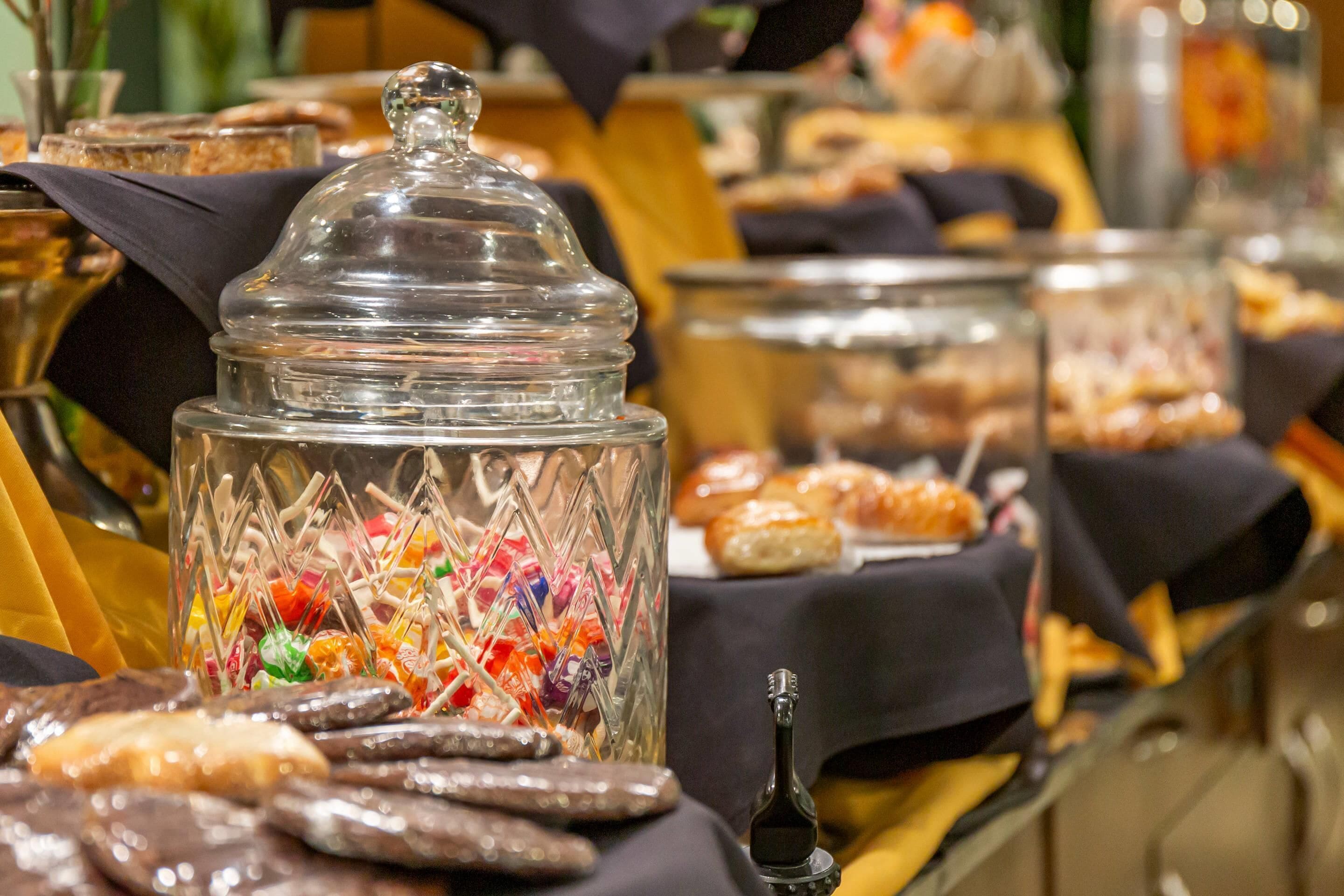 Buffet com diversos alimentos, incluindo um pote de vidro cheio de balas coloridas e uma pequena garrafa.