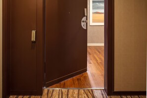 Entrada de um quarto de hotel com chão de madeira, porta marrom e vista para uma praia pela janela.