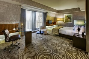 Quarto de hotel com cama, escrivaninha, poltrona, televisão e um quadro na parede.