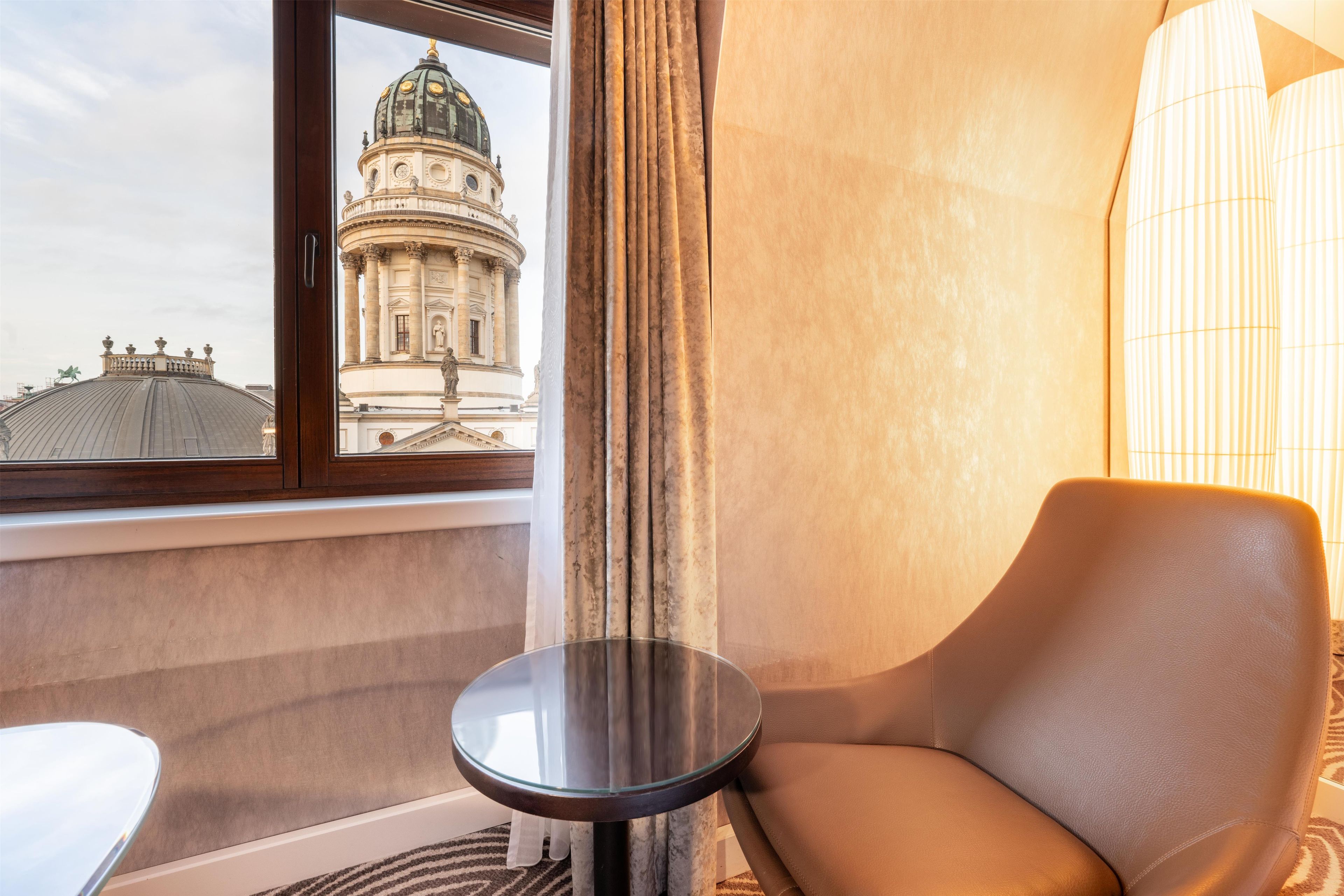 Quarto de hotel com vista para um edifício histórico através de uma janela, uma pequena mesa redonda e uma poltrona de couro.