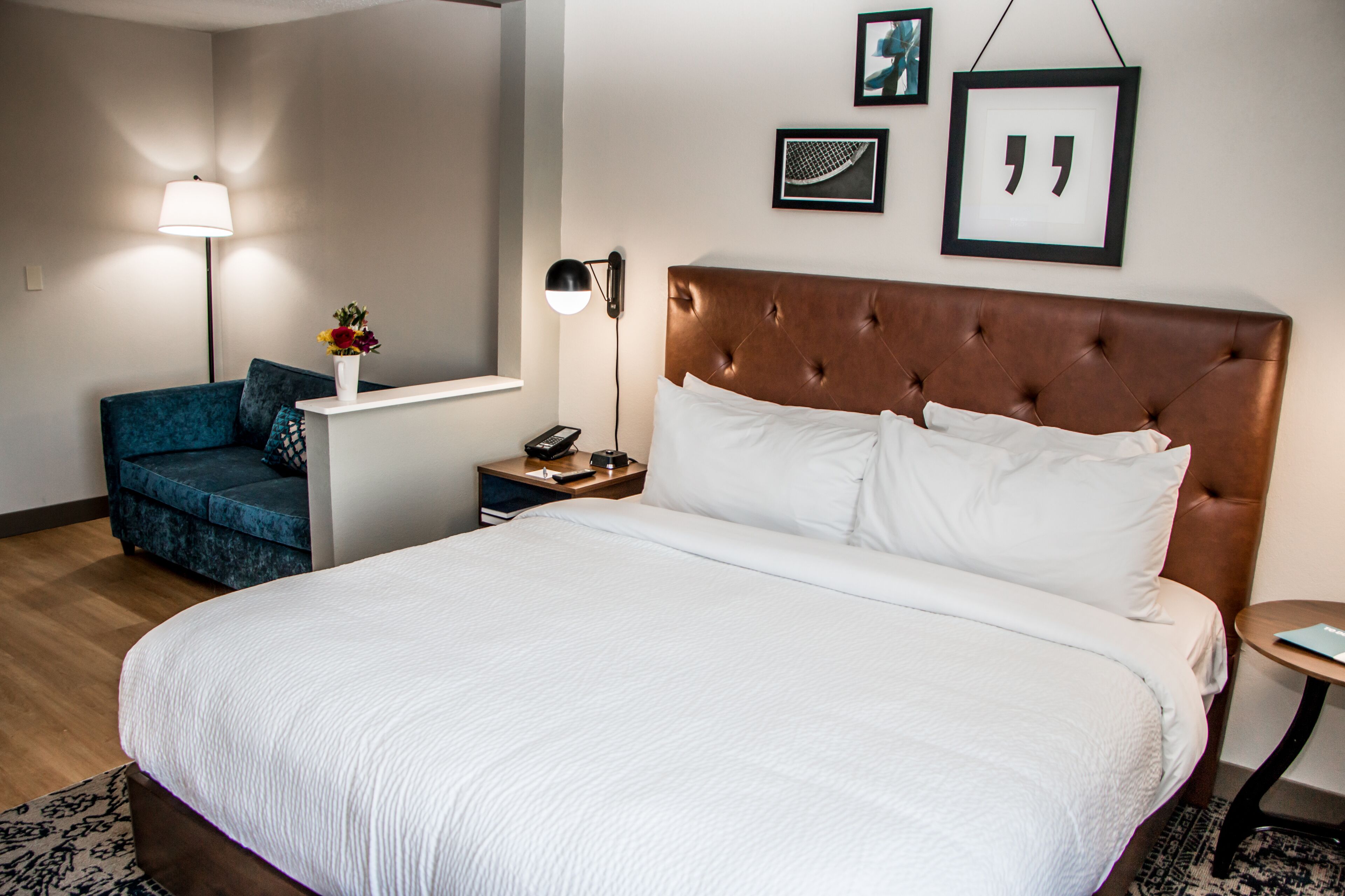 Quarto de hotel com uma cama grande, um sofá azul, uma mesa de cabeceira com abajur e quadros nas paredes.