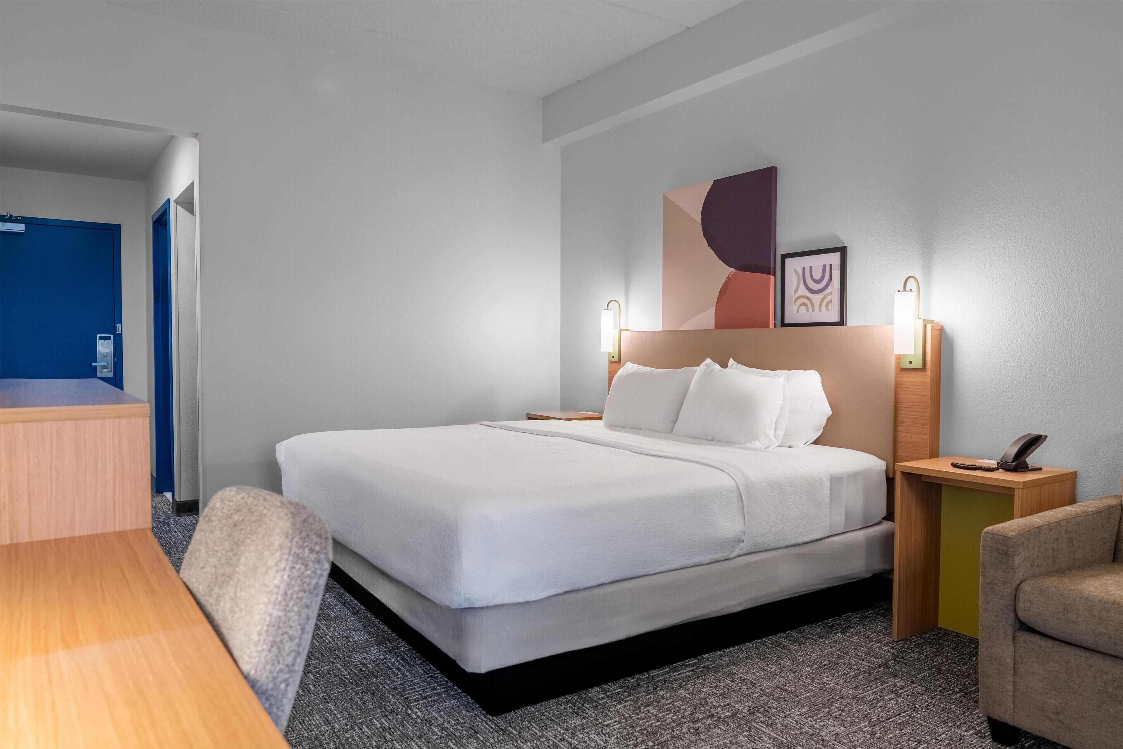 Quarto de hotel com uma cama, uma escrivaninha, uma cadeira e uma obra de arte na parede.