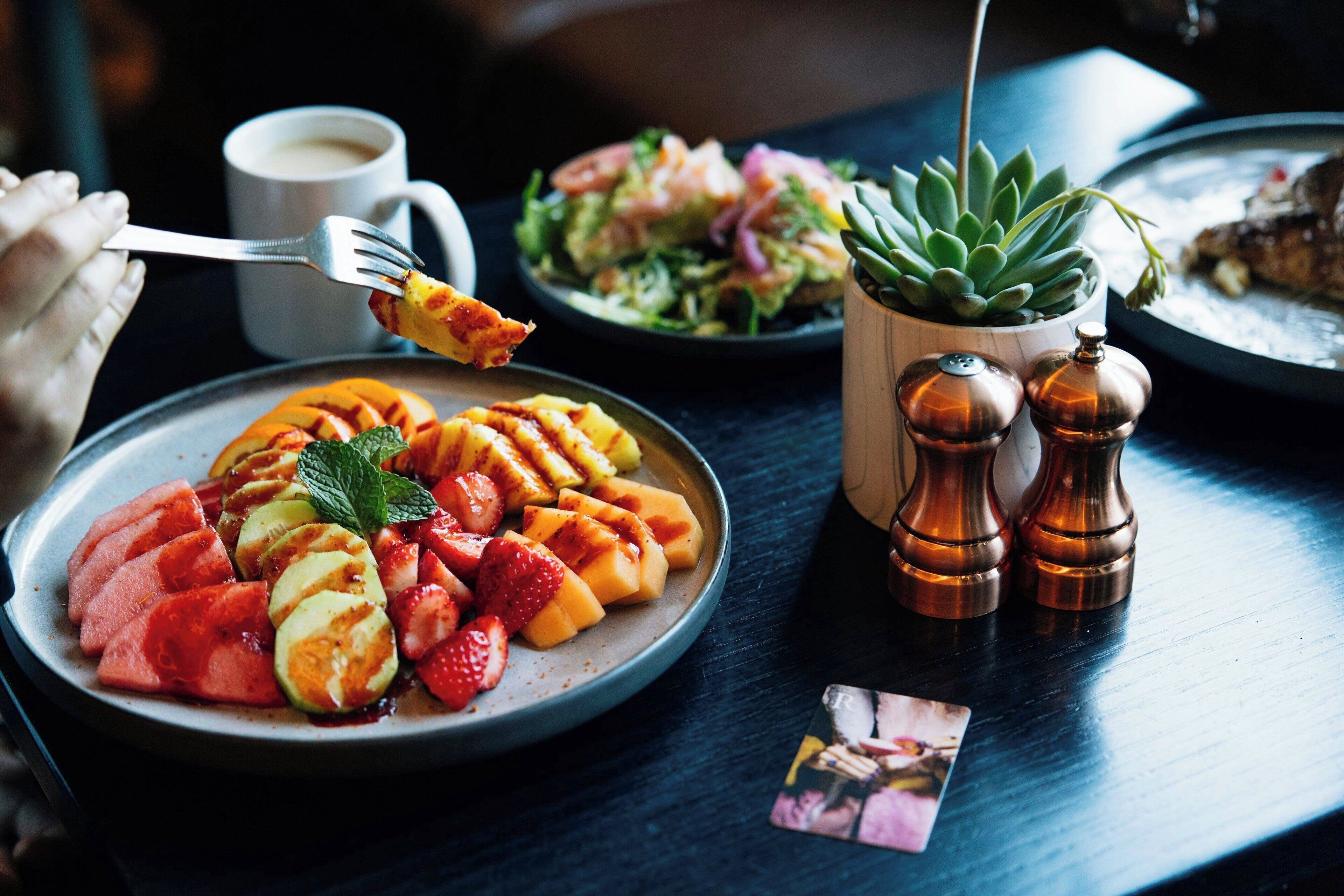 Uma mesa de jantar com uma salada de frutas, uma chávena de café e uma planta suculenta.