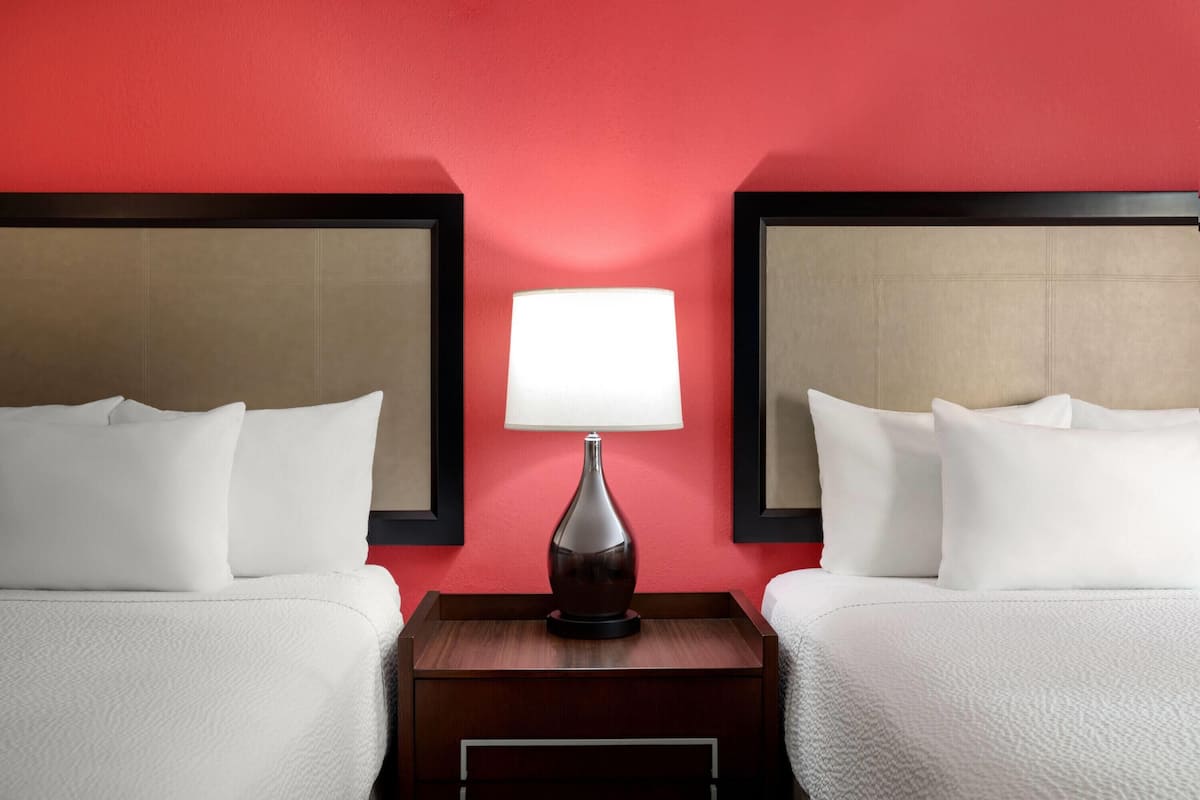 Quarto de hotel com duas camas, uma mesa de cabeceira com abajur e uma parede vermelha.