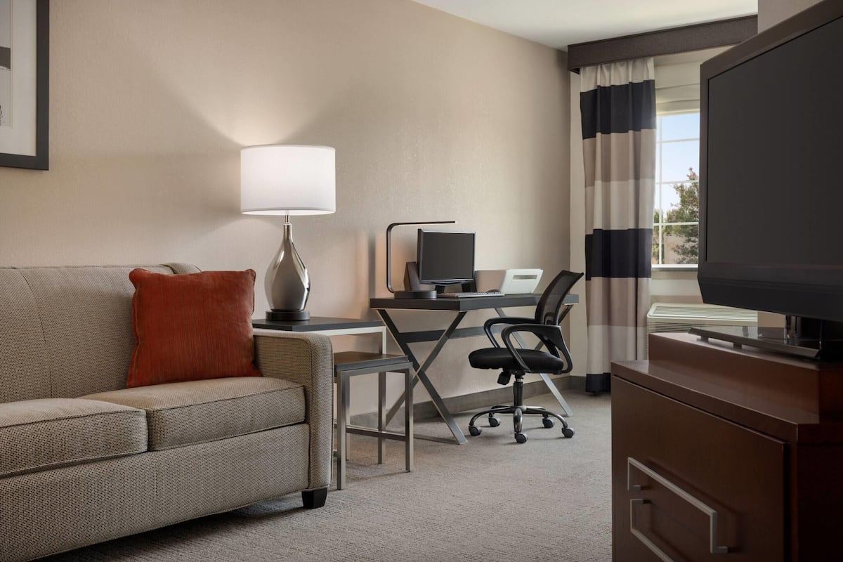 Quarto de hotel com sofá, escrivaninha, computador, cadeira, televisão e armário.