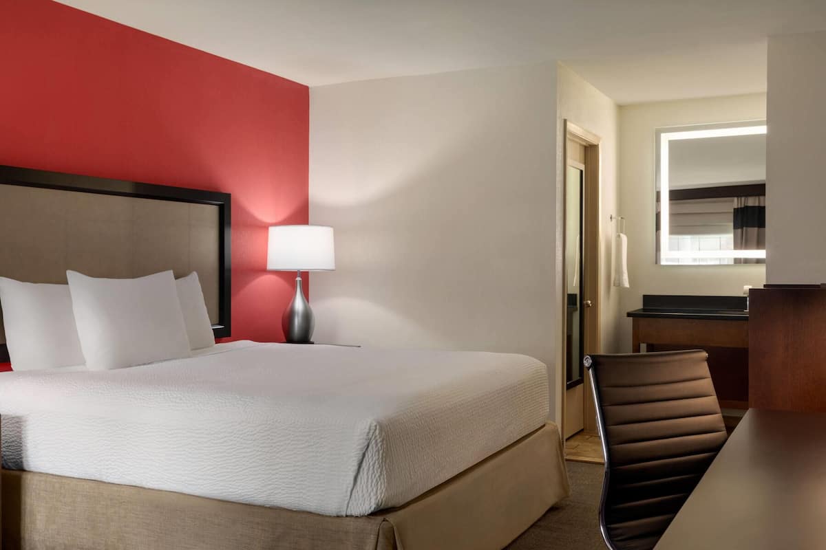 Quarto de hotel com cama, escrivaninha, cadeira, abajur e espelho.