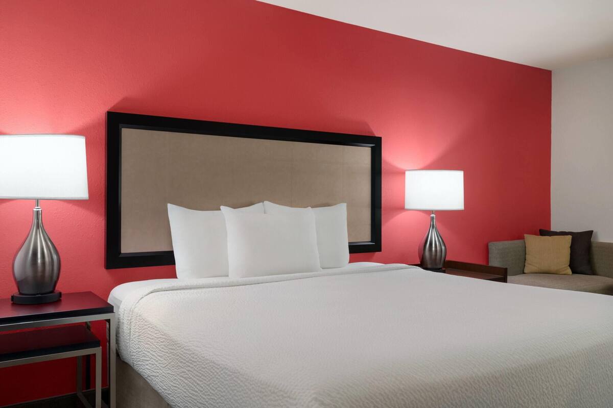 Quarto de hotel com uma cama grande, duas luminárias de cabeceira, uma parede com detalhes vermelhos e um sofá cinza.