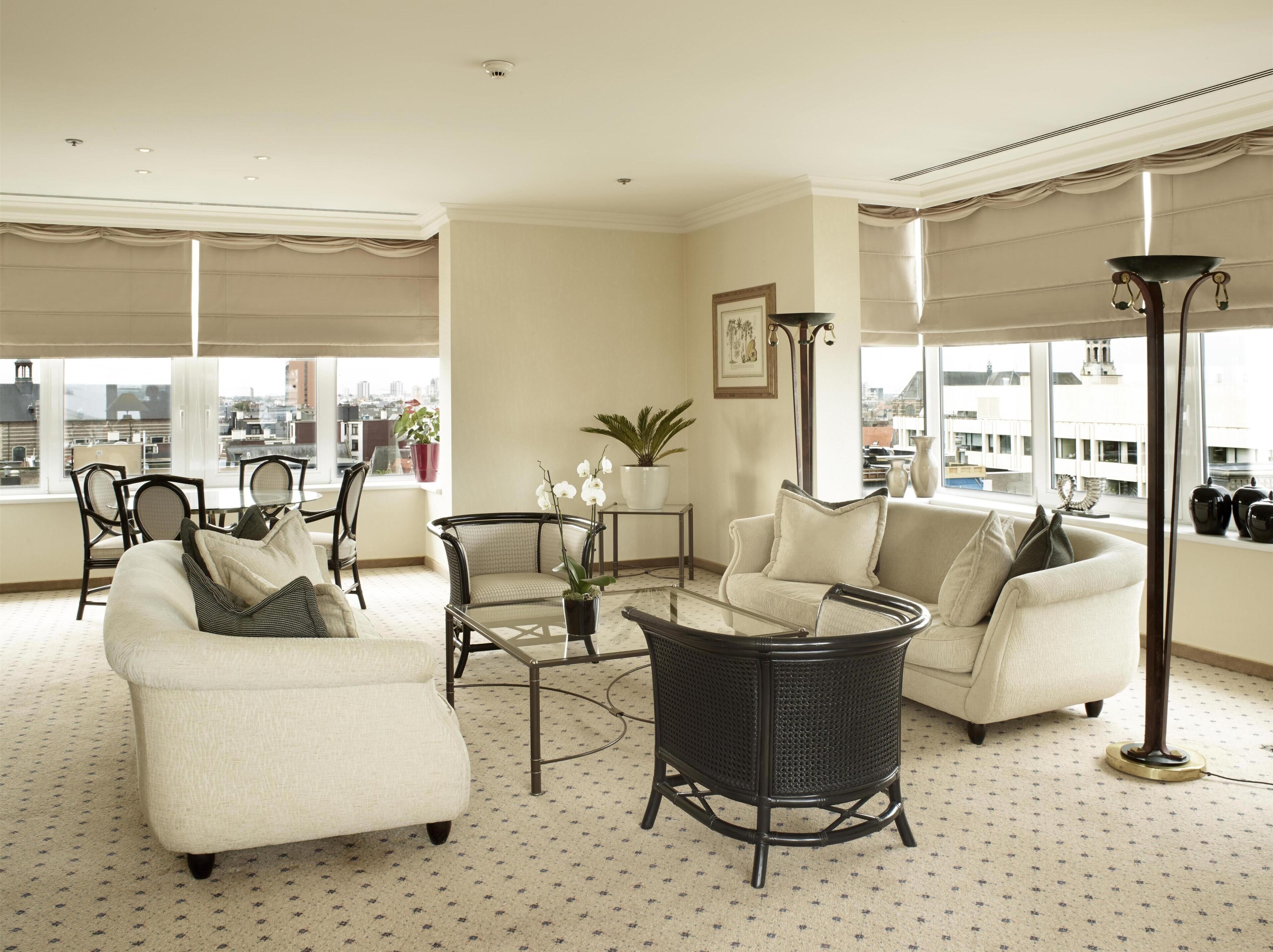 Uma sala de estar ampla com sofá, poltronas, mesa de centro e luminária de chão. Há plantas em vasos e vista para a cidade através de janelas grandes.
