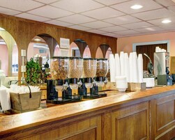 Um balcão de pequeno-almoço com vários dispensadores para cereais e granola, uma máquina de café e pilhas de copos de papel brancos.