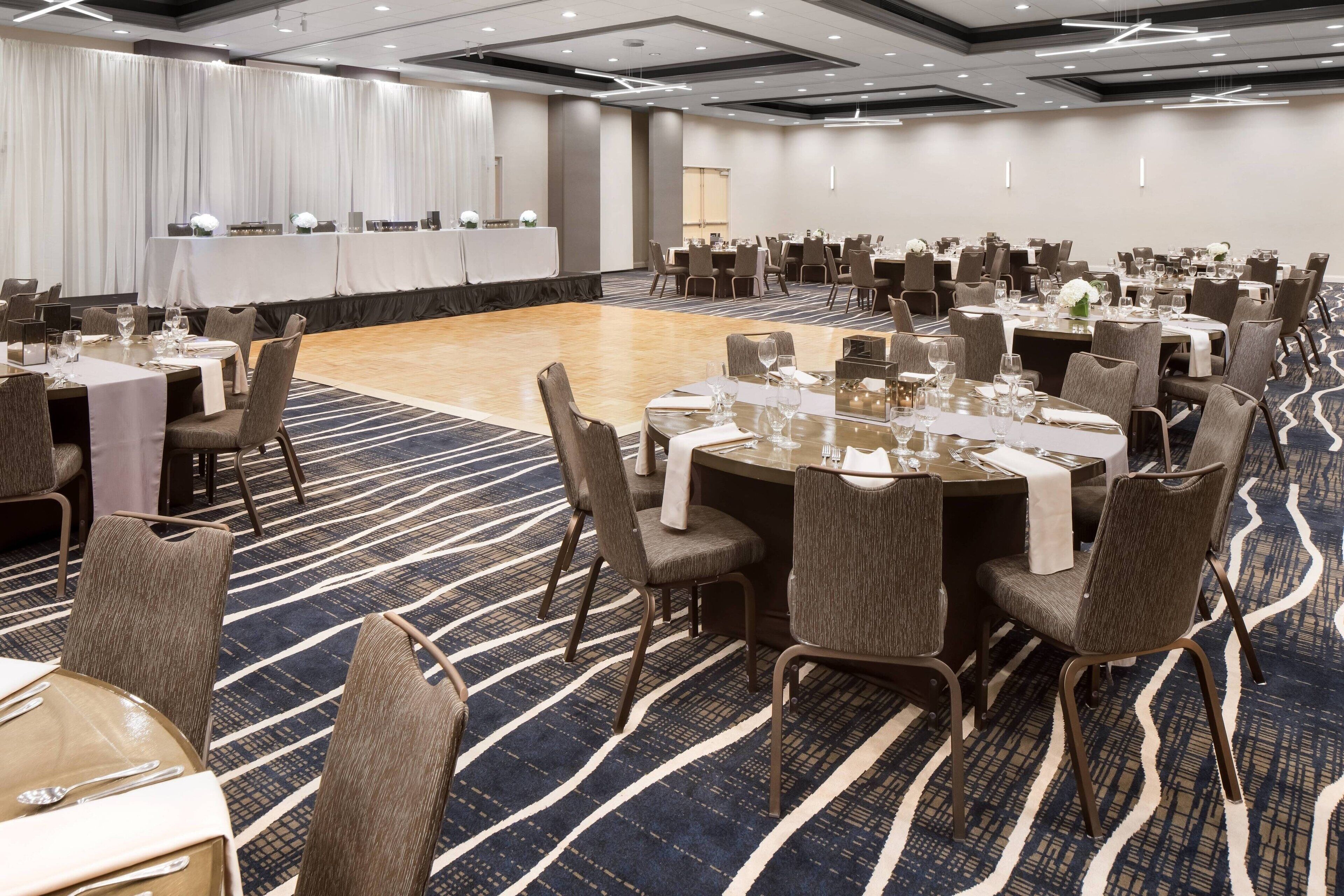 Um amplo salão de banquetes com mesas redondas e cadeiras, um palco com fundo branco e um tapete estampado.