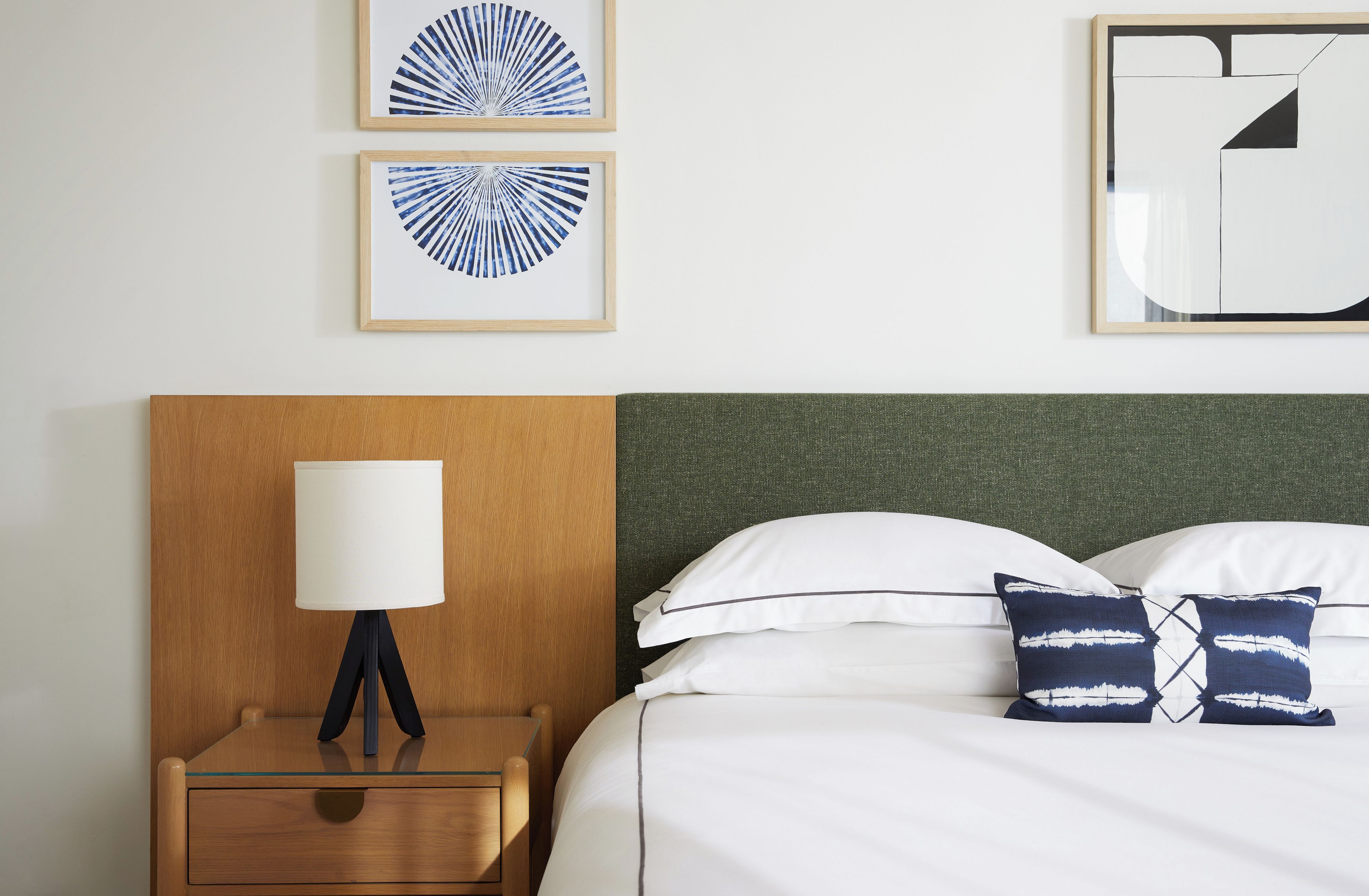 Cama bem arrumada com roupa de cama branca e dois travesseiros estampados em azul e branco, mesa de cabeceira de madeira com uma luminária, e duas obras de arte abstrata emolduradas na parede.