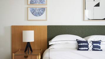 Cama bem arrumada com roupa de cama branca e dois travesseiros estampados em azul e branco, mesa de cabeceira de madeira com uma luminária, e duas obras de arte abstrata emolduradas na parede.