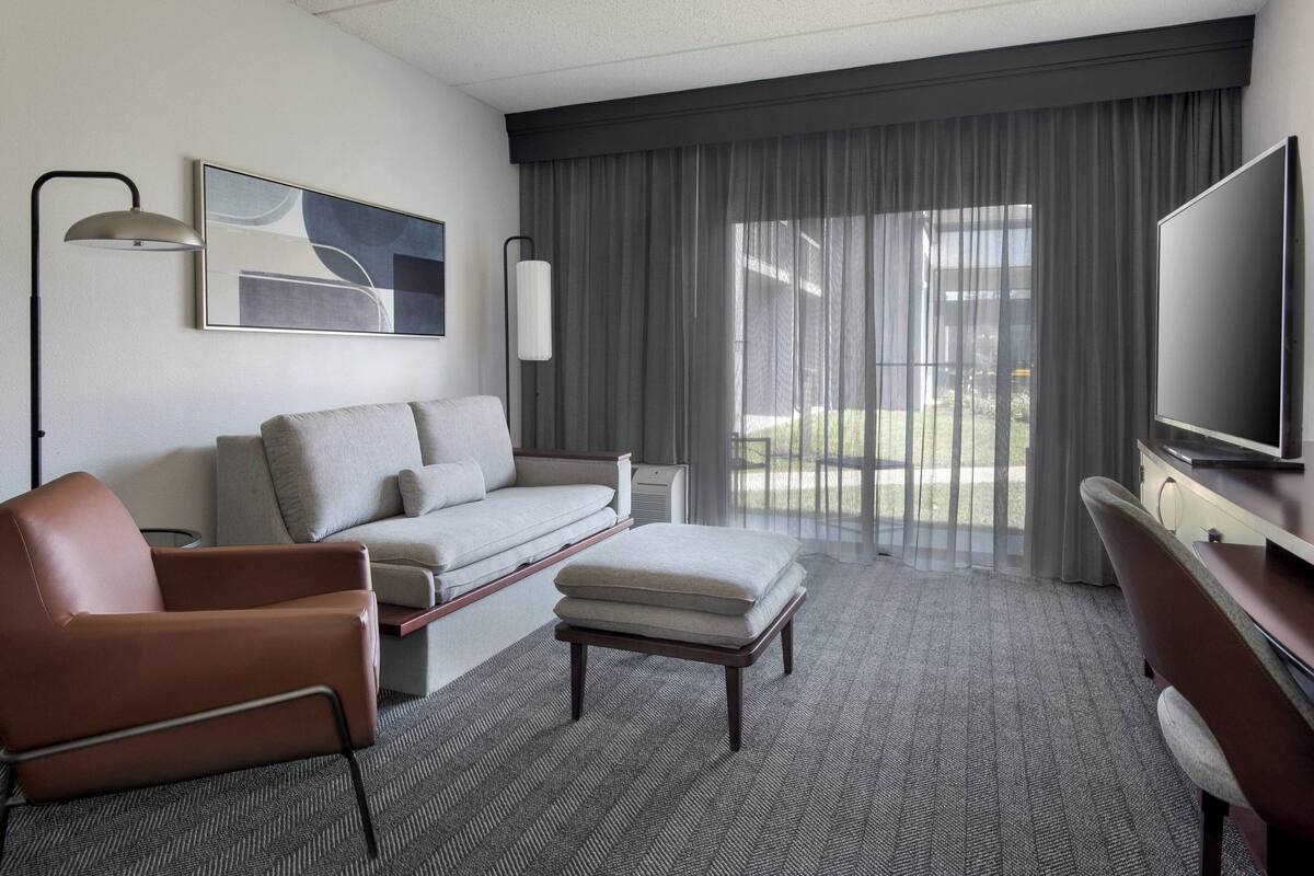 Quarto de hotel moderno com sofá, poltrona, banquinho, TV de tela plana e janelas amplas com cortinas leves.